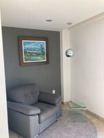 Uma sala de estar moderna com um sofá cinzento, um quadro emoldurado de uma paisagem, uma mesa de centro de vidro e um candeeiro fixado ao teto.
