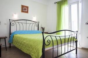 Uma cama com colcha verde e azul, uma pequena mesa de cabeceira, uma luminária na parede, um quadro emoldurado e uma cortina verde.