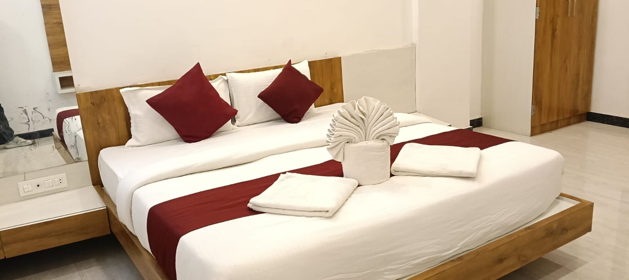 Quarto de hotel moderno com cabeceira de madeira, roupa de cama branca, detalhes em vermelho e um vaso decorativo.