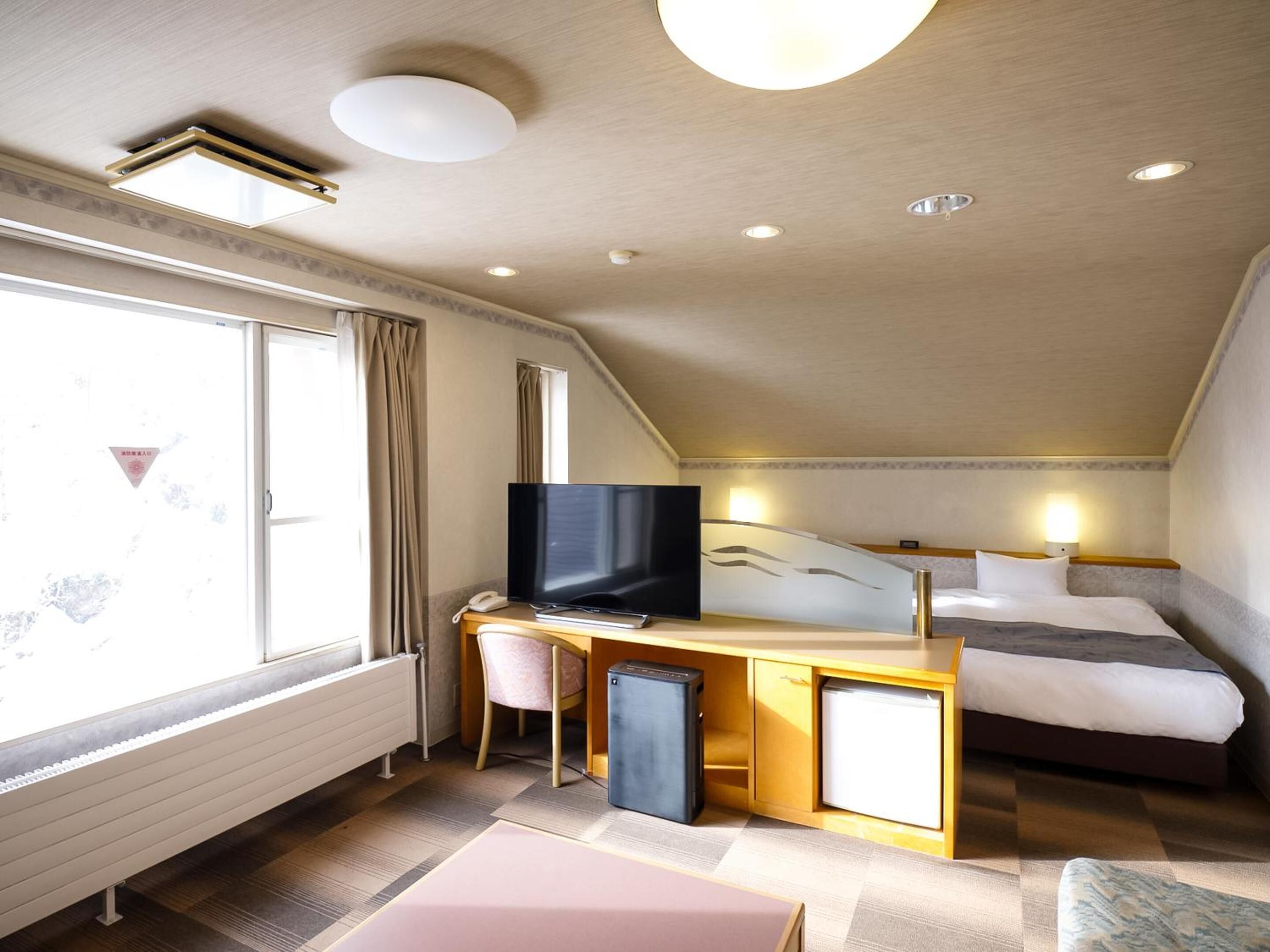 Quarto de hotel com cama, escrivaninha com televisão, cadeira, mesinha de centro e janela com cortinas.
