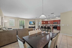 Uma cozinha moderna com armários escuros, uma sala de jantar com uma mesa grande e uma sala de estar com sofá e poltronas.