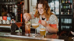 Um bartender preparando bebidas em um bar, com vários frascos e copos à mostra.