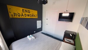 Quarto de hotel com uma cama, uma TV fixada na parede e um aviso que diz 'FIM DA OBRA'.