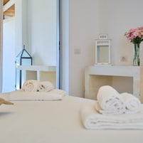 Quarto de hotel com cama, mesa de cabeceira com espelho, vaso de flores e porta-toalhas.