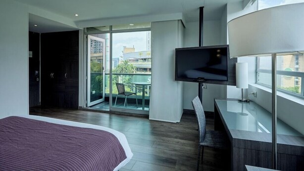Quarto de hotel moderno com uma cama grande, uma escrivaninha, uma TV e uma varanda com vista para edifícios.