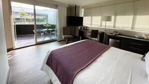 Quarto de hotel moderno com uma cama grande, uma escrivaninha, uma cadeira, uma televisão e uma porta de vidro deslizante para o exterior.