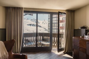 Quarto com uma janela grande, uma escrivaninha de madeira, uma cadeira e vista para montanhas cobertas de neve.