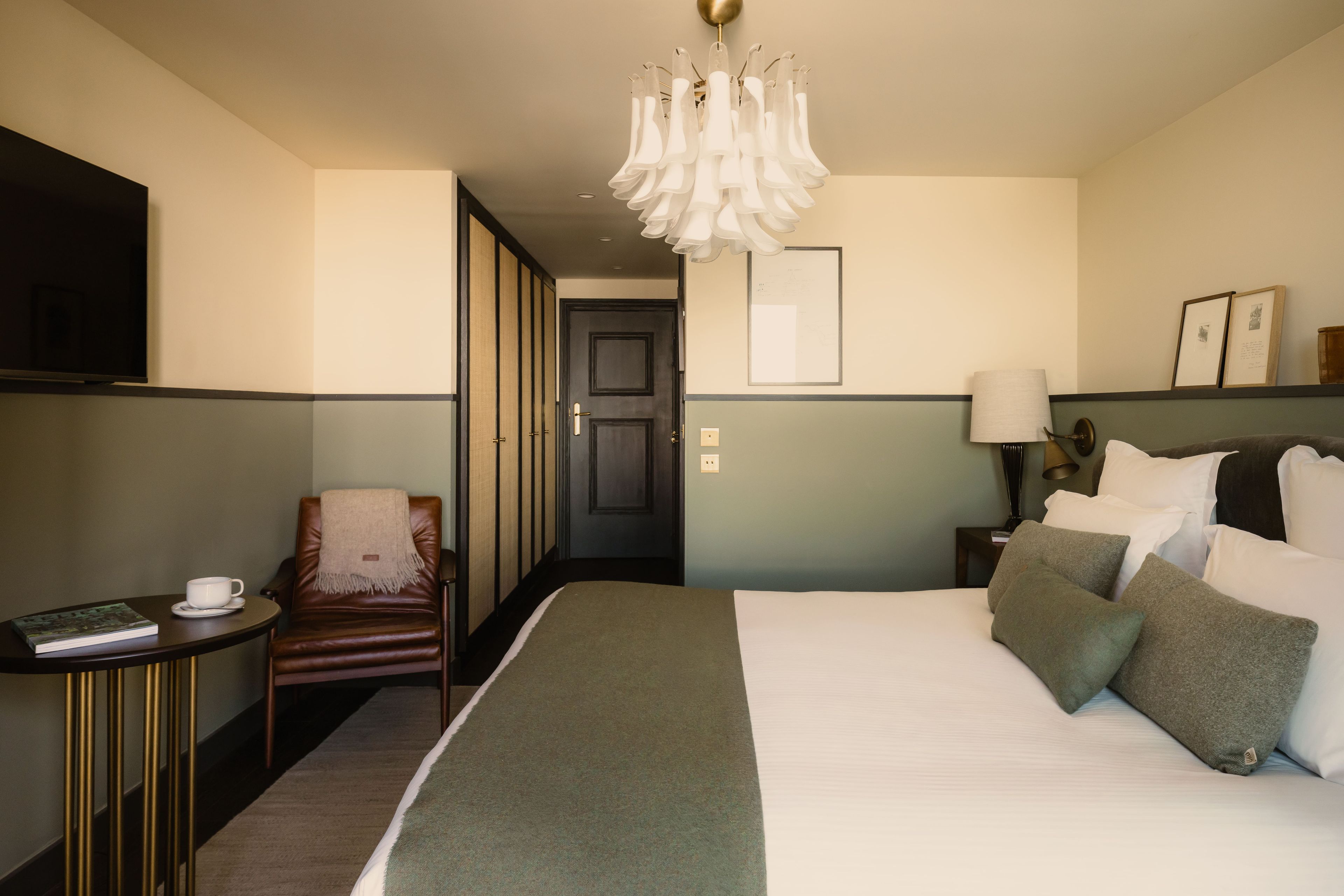 Quarto de hotel com uma cama, uma cadeira, uma mesa com um livro, uma luminária e uma televisão.