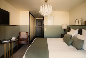 Quarto de hotel com uma cama, uma cadeira, uma mesa com um livro, uma luminária e uma televisão.