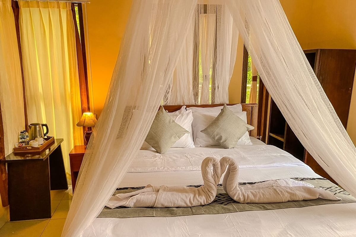 Uma cama com dossel e cortinas, uma mesa de cabeceira com abajur e vista para o exterior pela janela.