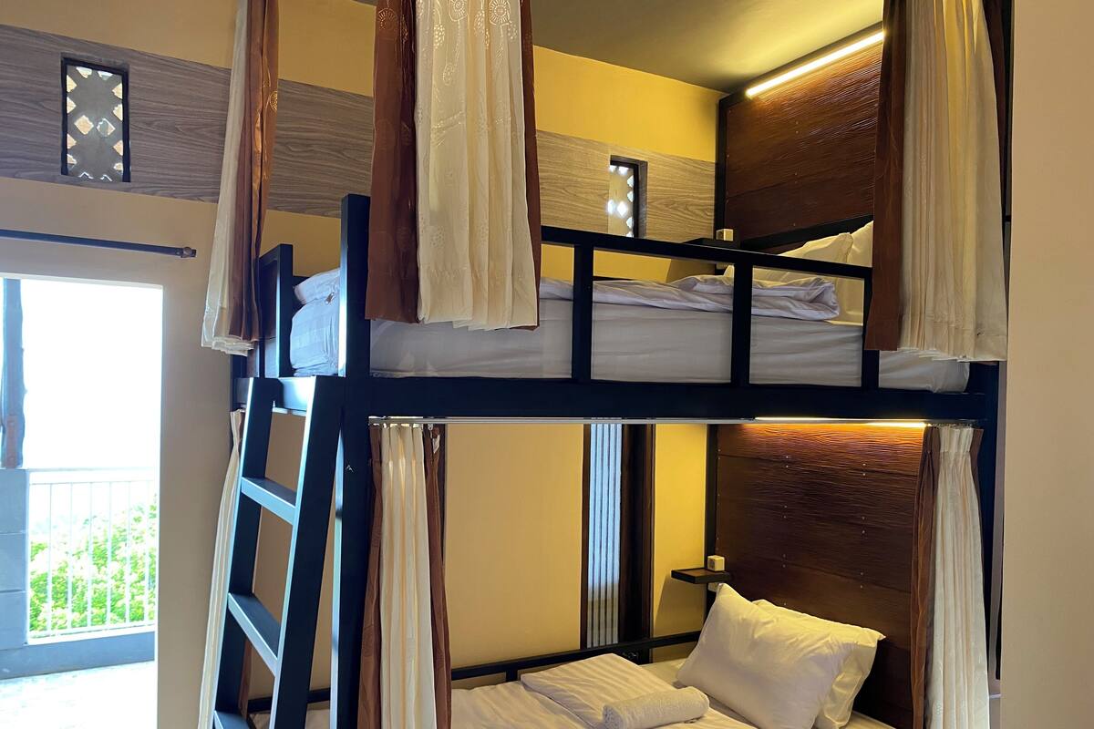 Quarto com beliche, prateleira pequena e janela com vista para o verde.
