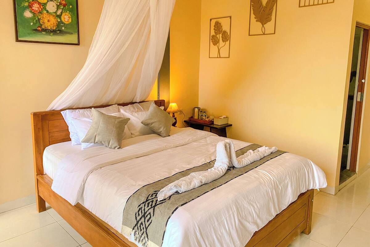Uma cama bem arrumada com dossel, uma mesa de cabeceira com abajur, e um quadro de flores na parede.