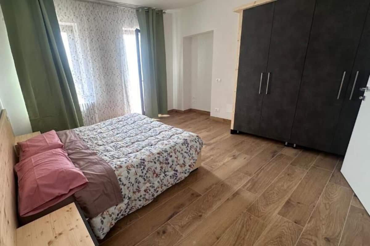 Quarto com cama de madeira, guarda-roupa escuro e janela com cortinas verdes.