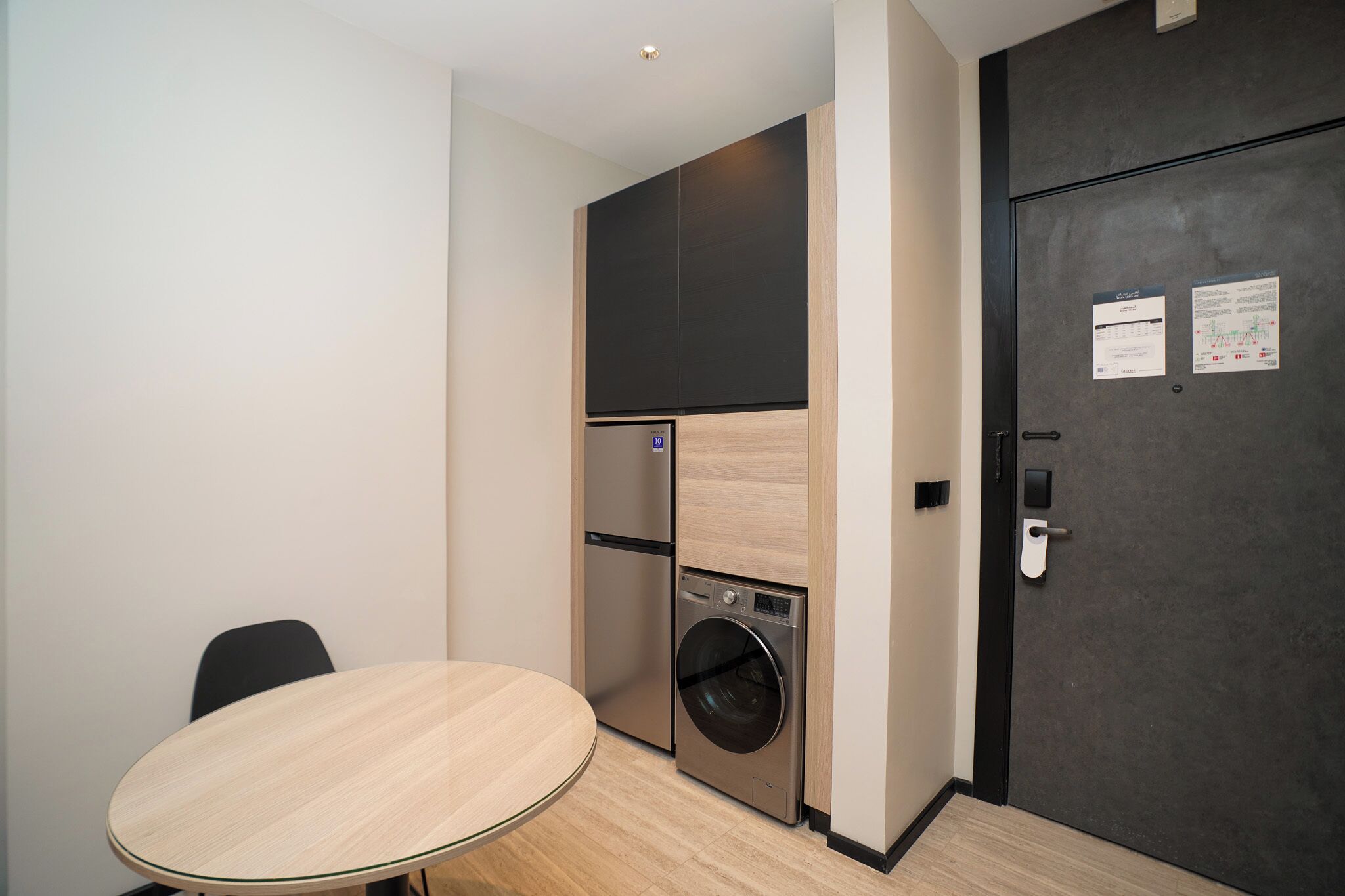 Quarto de hotel compacto com uma pequena mesa, máquina de lavar, frigorífico e porta com fechadura e leitor de cartão.