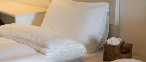 Cama bem arrumada com lençóis e almofadas brancas, mesa de cabeceira com caixa de lenços de papel com a etiqueta "H. suite".