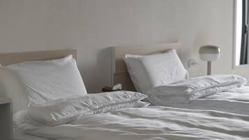Cama bem arrumada com roupa de cama e almofadas brancas, mesa de cabeceira com abajur e janela com cortinas.