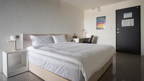 Quarto moderno com uma cama grande, mesa de cabeceira com luminária, escrivaninha e porta com pôster.