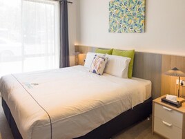 Quarto de hotel com uma cama grande, uma mesa de cabeceira com abajur, uma gaveta pequena e uma obra de arte pendurada na parede.