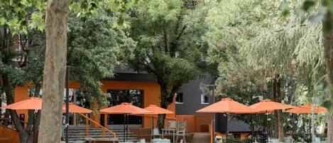 Uma área externa com mesas e cadeiras, com guarda-sóis laranja, cadeiras brancas e um lance de escadas que leva a um edifício com fachada escura.