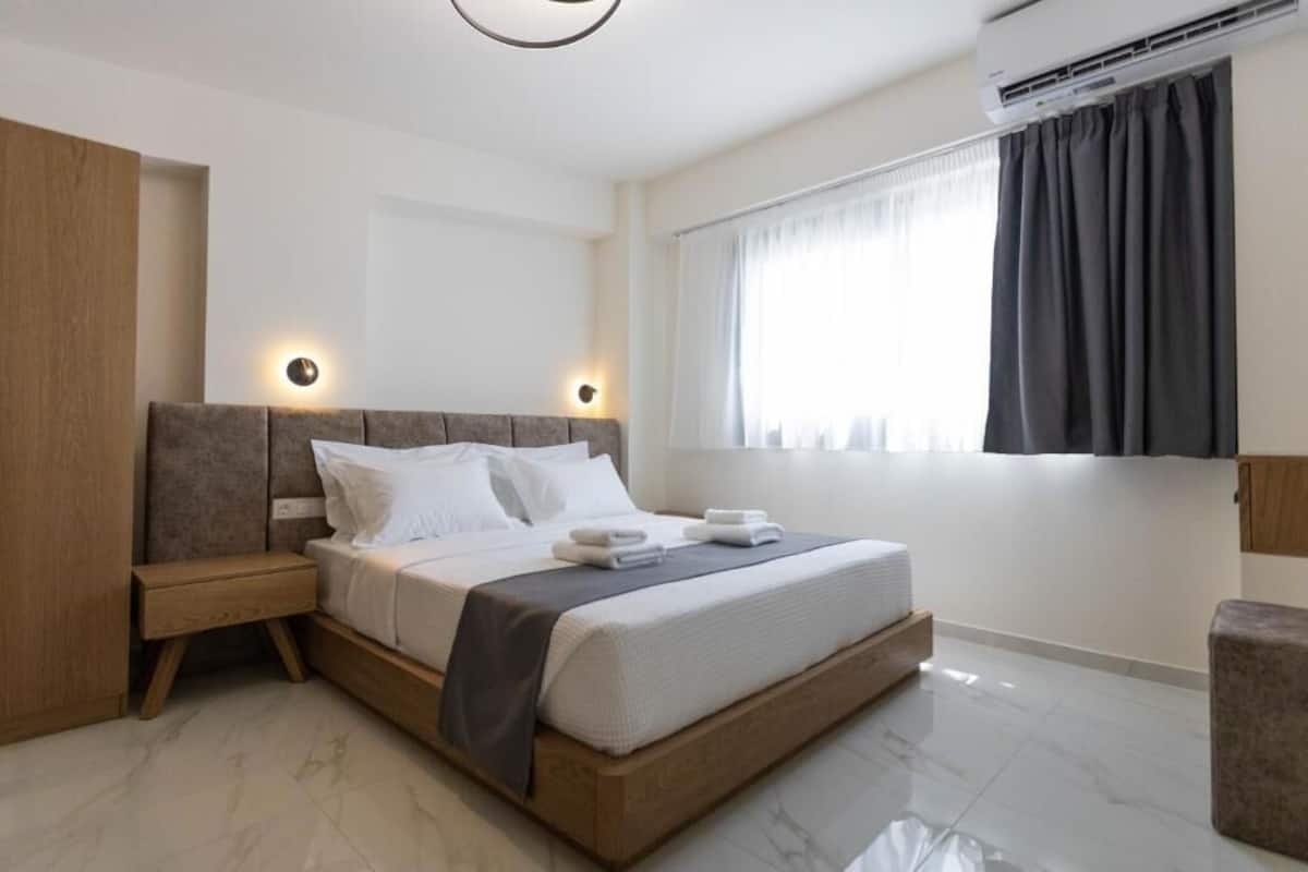 Quarto de hotel moderno com uma cama grande, mesas de cabeceira, um guarda-roupa e uma janela com cortinas.