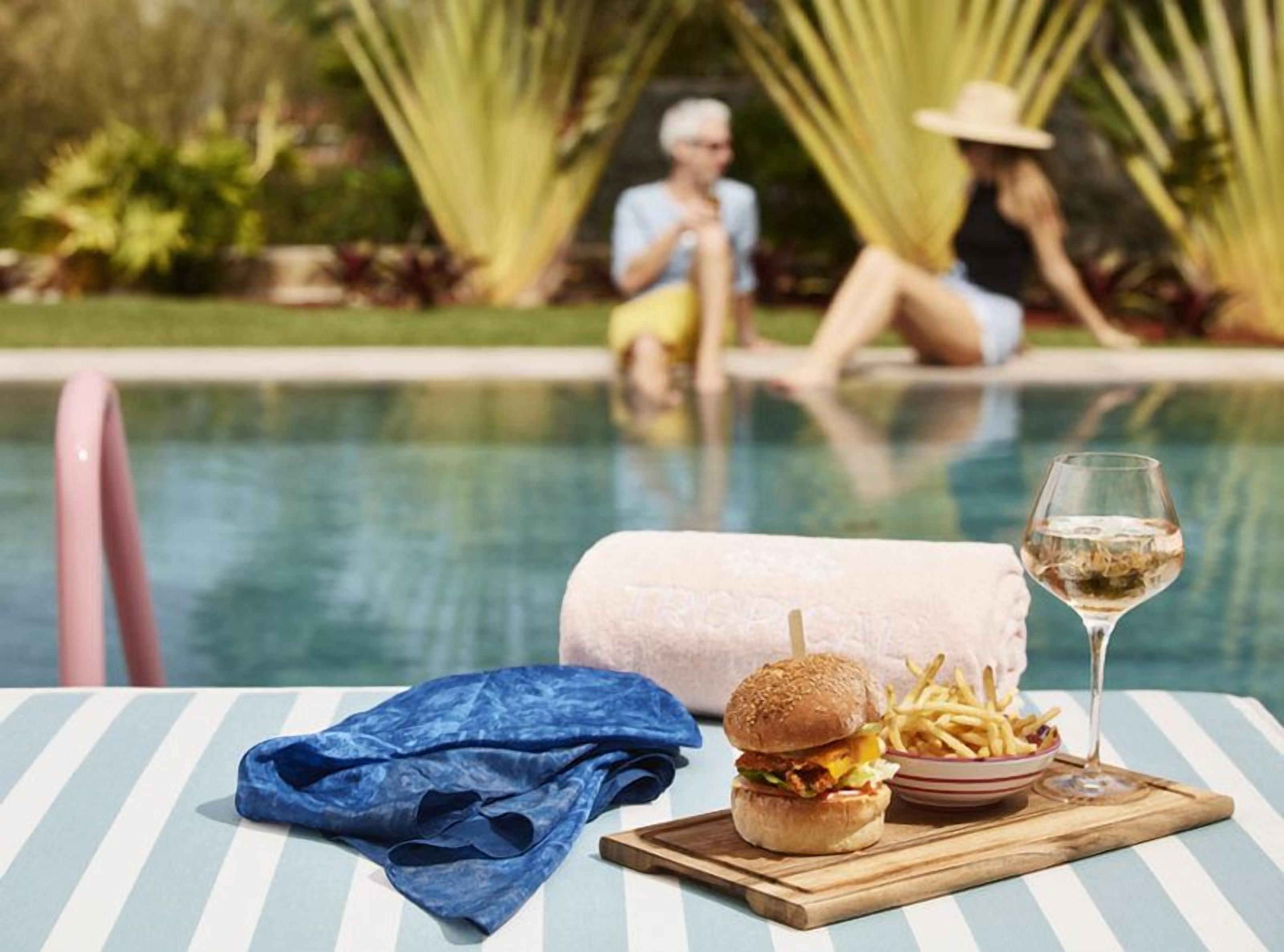 Ambiente à beira da piscina com mesa, servindo um hambúrguer, batatas fritas e um copo de vinho.