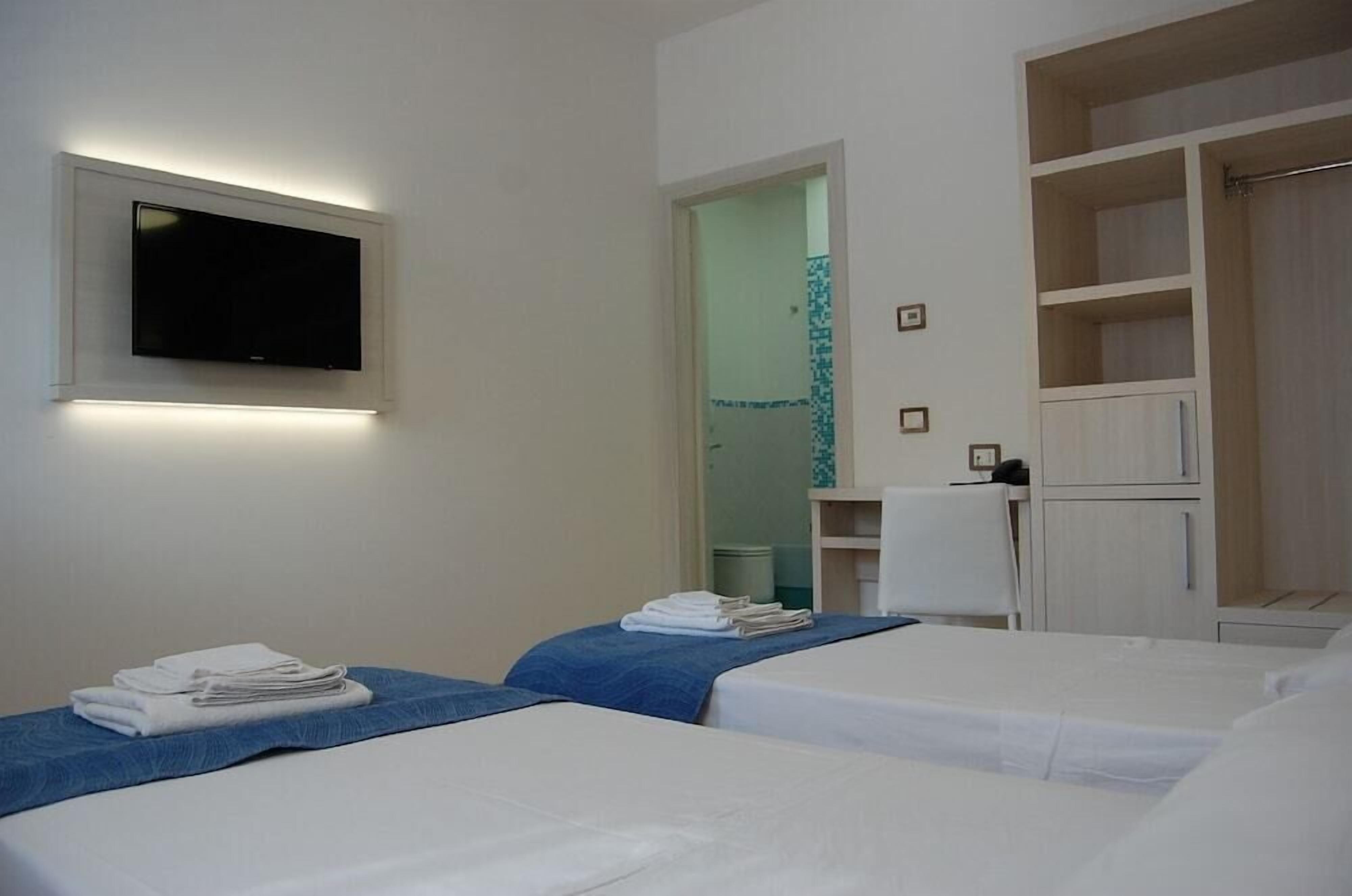 Quarto de hotel com duas camas, televisão na parede, guarda-roupa e pequena mesa de trabalho com cadeira.