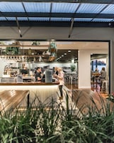 Um café moderno com balcão de madeira, prateleiras com plantas e área de estar com mesas e cadeiras.