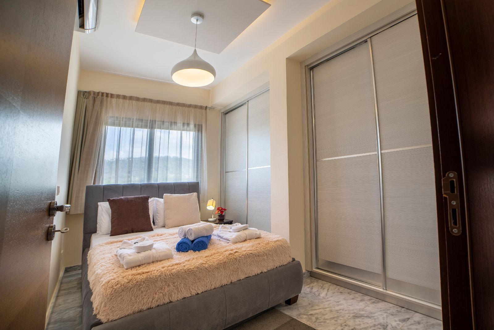 Quarto de hotel com cama, mesa de cabeceira, luminária, janela com cortinas e porta de correr.