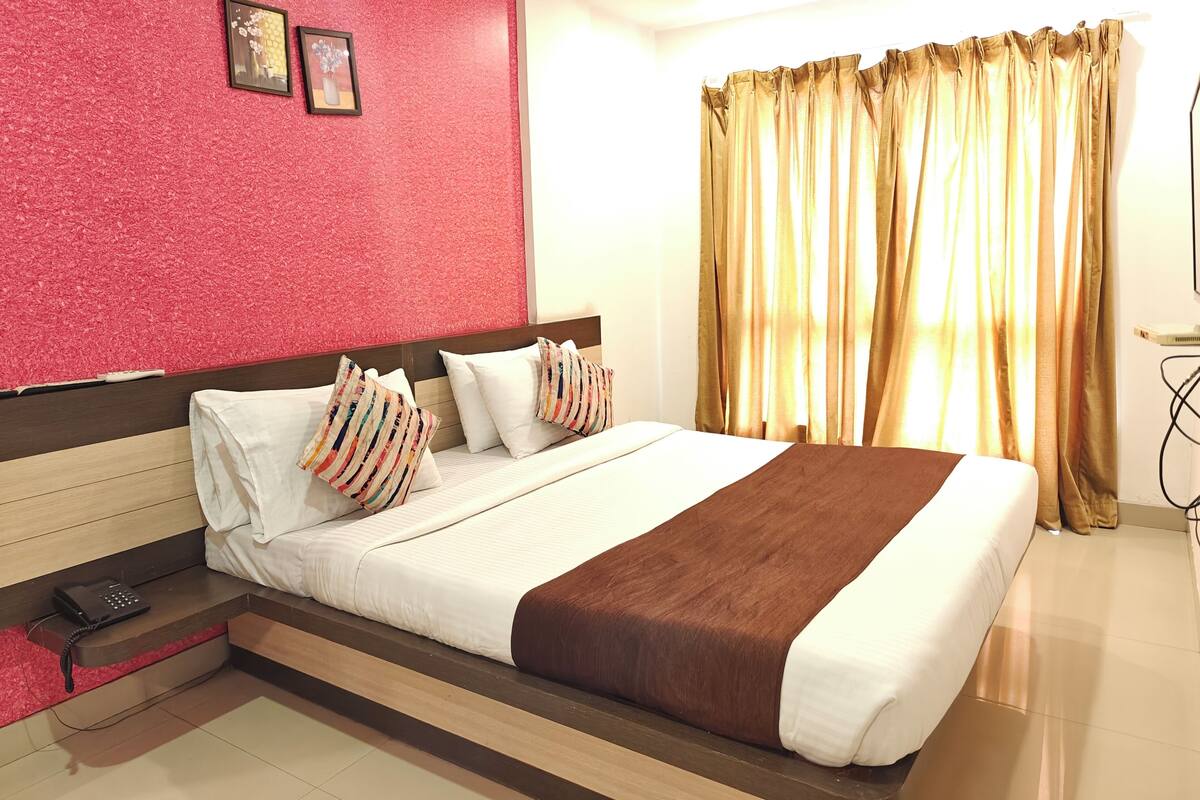 Quarto de hotel com uma cama grande, parede vermelha com textura e uma janela com cortinas douradas.
