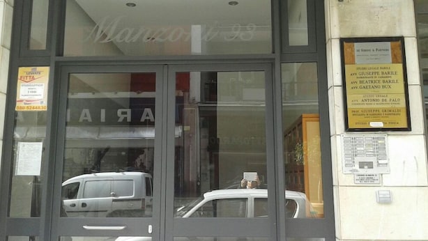 Entrada de um edifício com portas de vidro, uma placa de identificação e o reflexo de um carro.