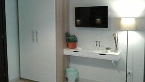 Quarto com guarda-roupa, TV fixada na parede, mesa de cabeceira com abajur e um pequeno cesto de lixo.