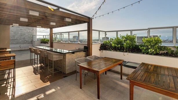 Um pátio externo com mesa de madeira, cadeiras, área de bar e vista para a cidade.