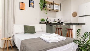 Quarto com cama, uma mesinha com um livro e óculos, e área de cozinha com micro-ondas e geladeira.