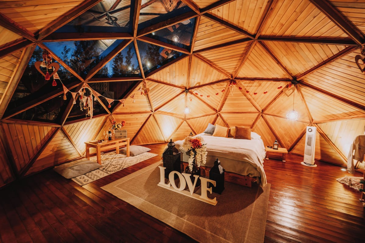 Uma tenda de madeira aconchegante em formato triangular, com uma cama, uma mesinha e um letreiro com a palavra 'LOVE' no centro.