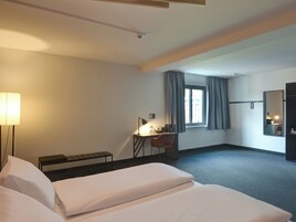 Quarto de hotel com cama, escrivaninha, cadeira, luminária e janela com cortinas.