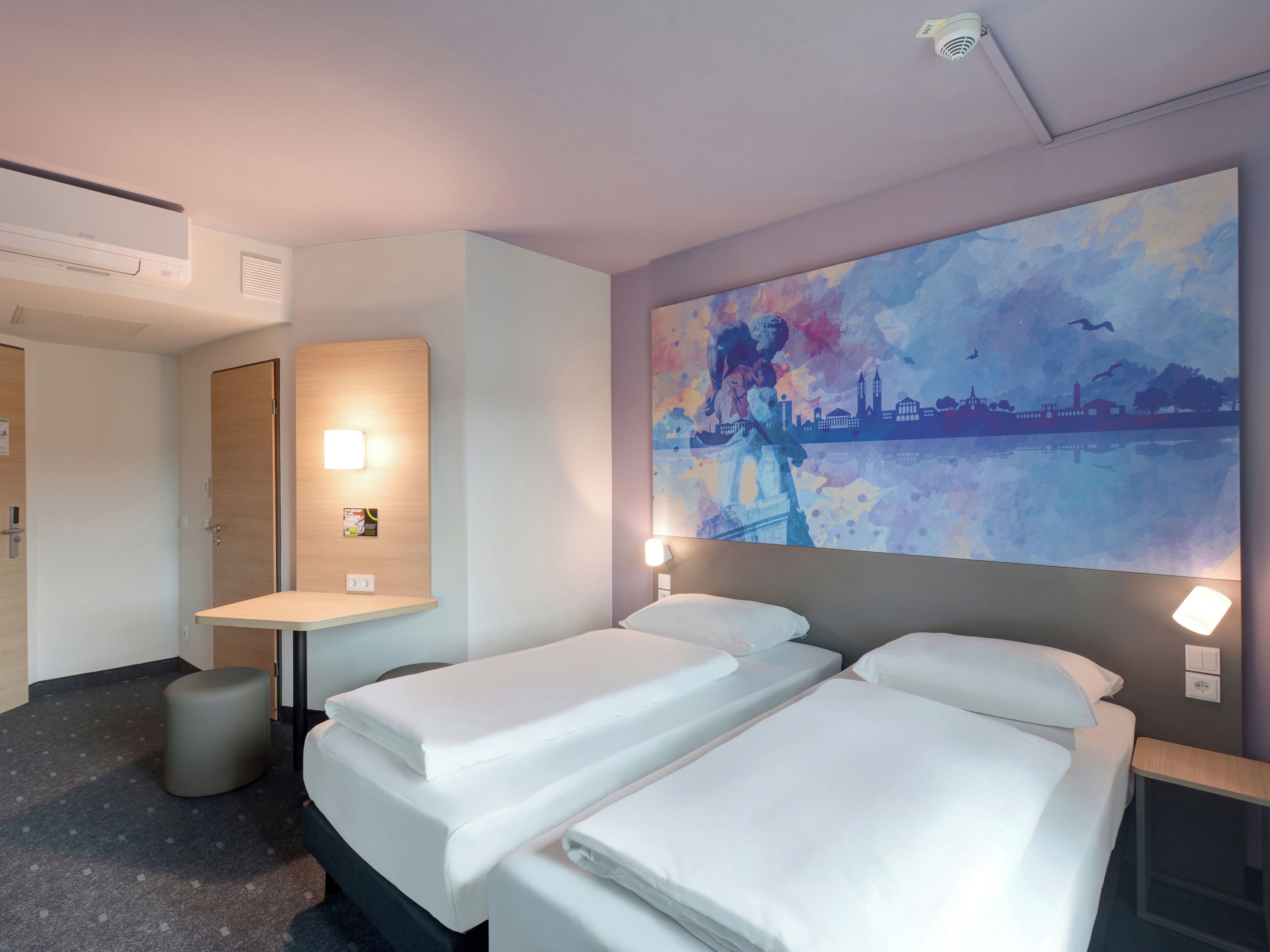 Quarto de hotel com duas camas, uma escrivaninha, uma cadeira, ar condicionado fixado na parede e uma grande pintura de uma paisagem urbana.