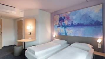 Quarto de hotel com duas camas, uma escrivaninha, uma cadeira, ar condicionado fixado na parede e uma grande pintura de uma paisagem urbana.