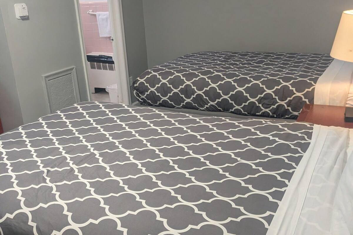 Quarto de hotel com duas camas, cada uma com colcha estampada. Há uma mesa de cabeceira com abajur, e uma porta que dá para um banheiro com porta-toalhas.