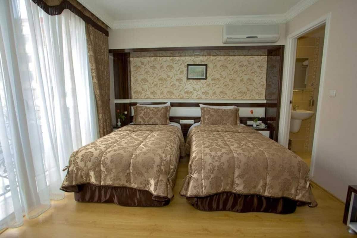 Quarto com cama de casal, piso de madeira, janela grande com cortinas, ar condicionado fixado na parede e banheiro visível por uma porta aberta.