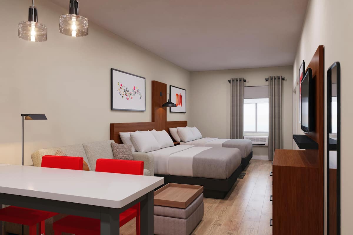 Quarto de hotel com duas camas, uma mesa de jantar, cadeiras vermelhas, televisão e obras de arte nas paredes.