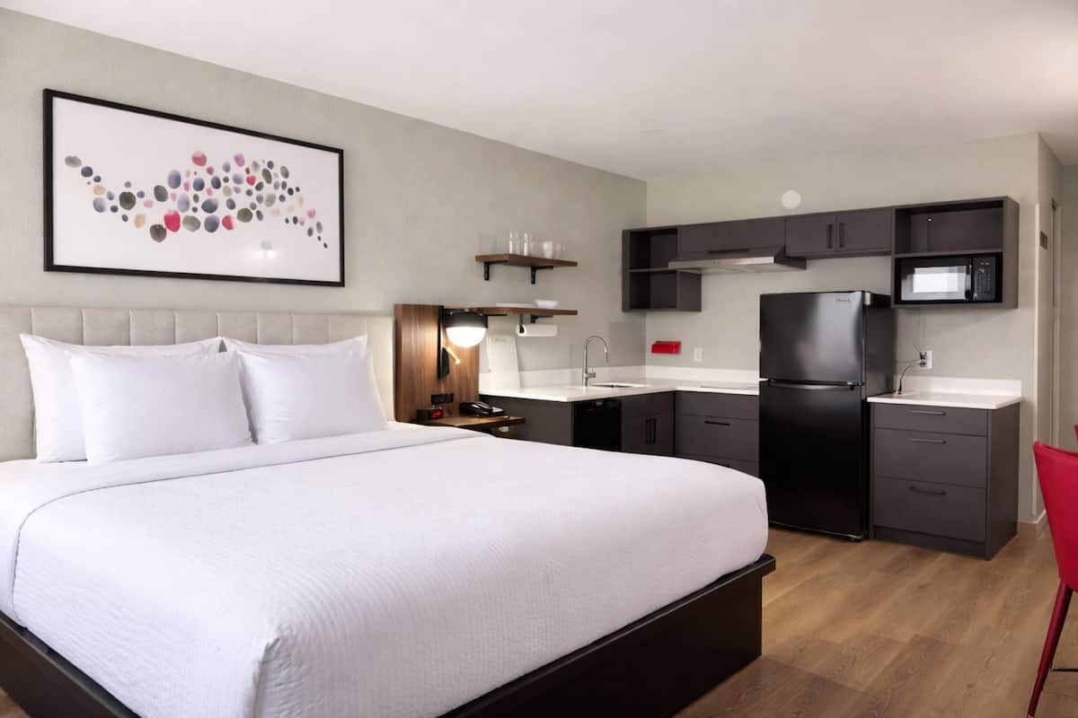 Quarto de hotel moderno com cama grande, cozinha compacta, micro-ondas e uma cadeira vermelha.