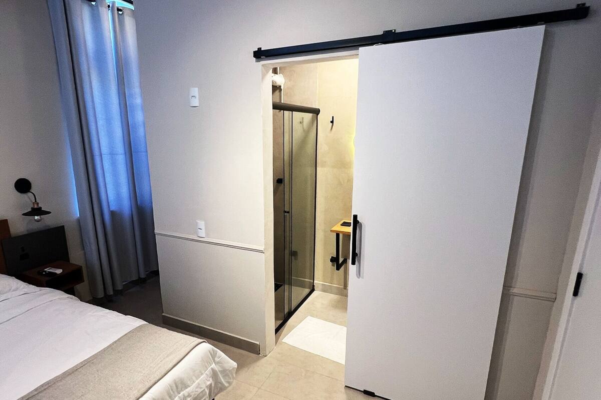 Quarto de hotel com cama, mesa de cabeceira, escrivaninha, cadeira, armário e banheiro com chuveiro e vaso sanitário.