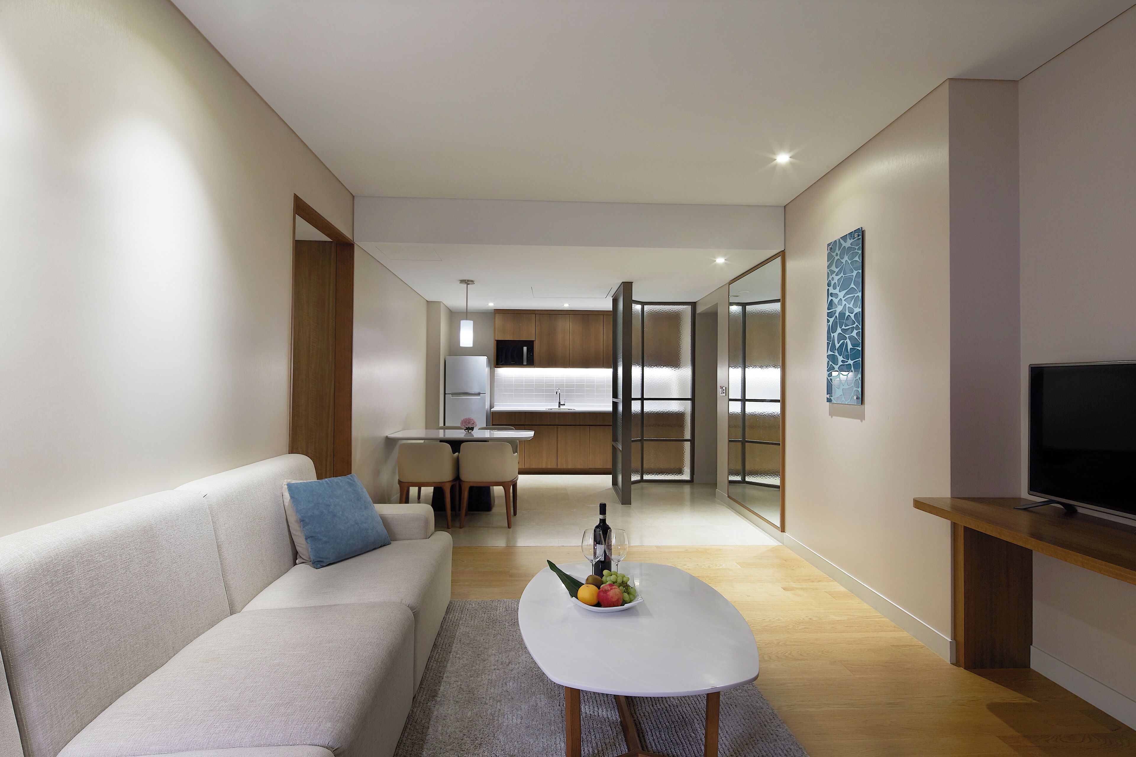 Uma sala de estar moderna com um sofá, uma mesa de centro com frutas, uma televisão e uma cozinha ao fundo.
