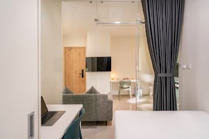 Quarto de hotel moderno com cama, escrivaninha com laptop, sofá, TV fixada na parede e porta de vidro que dá para o banheiro.