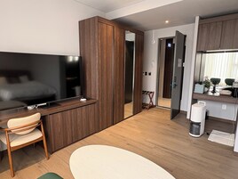 Quarto de hotel moderno com uma televisão de tela plana grande, móveis de madeira, uma pequena escrivaninha e uma cozinha compacta.
