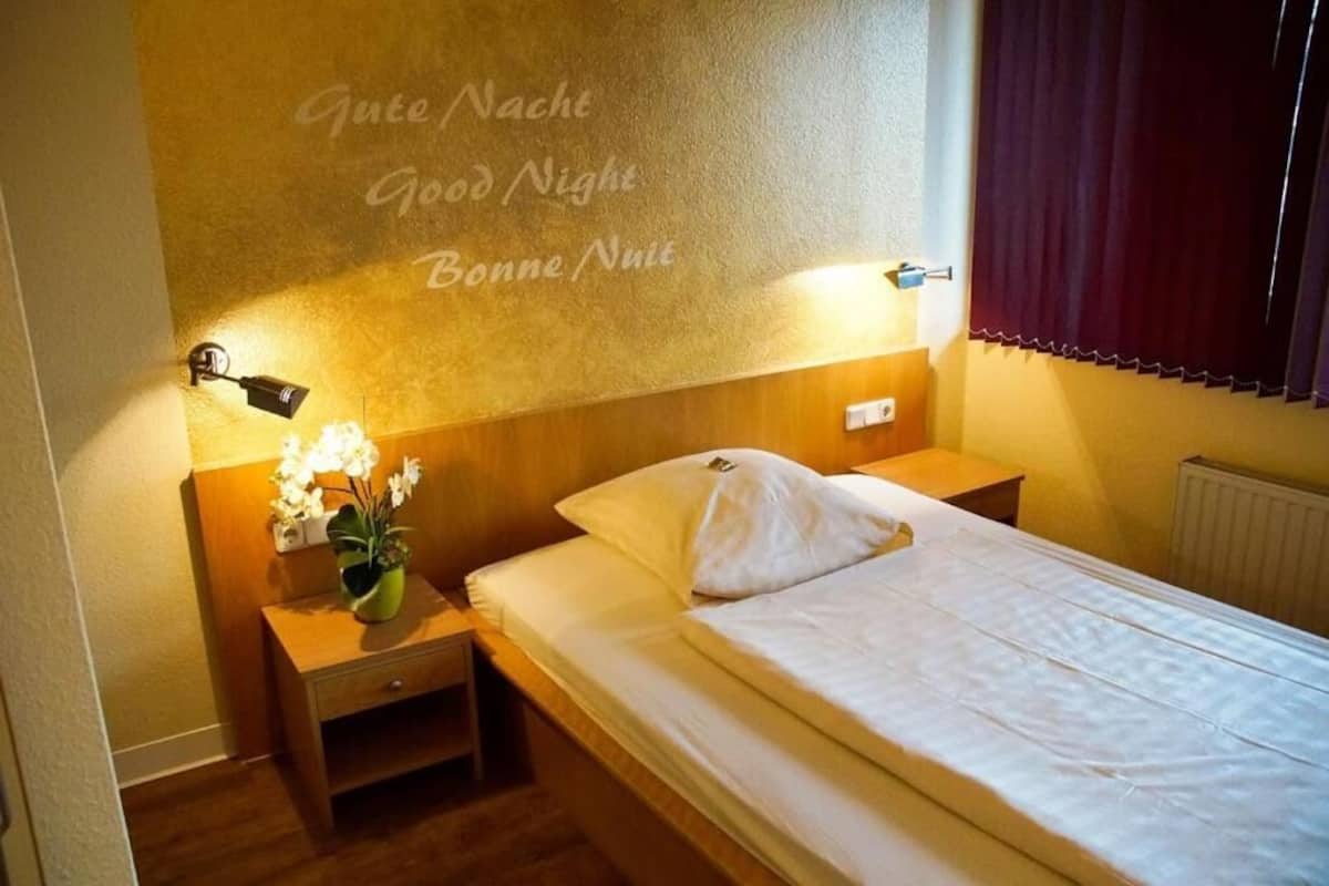 Quarto de hotel com cama, mesas de cabeceira, um criado-mudo com abajur e parede com a mensagem 'Boa Noite' em vários idiomas.