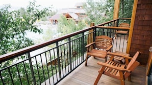 Um deck de madeira com guarda-corpo, duas cadeiras de madeira e vista para edifícios e árvores ao fundo.