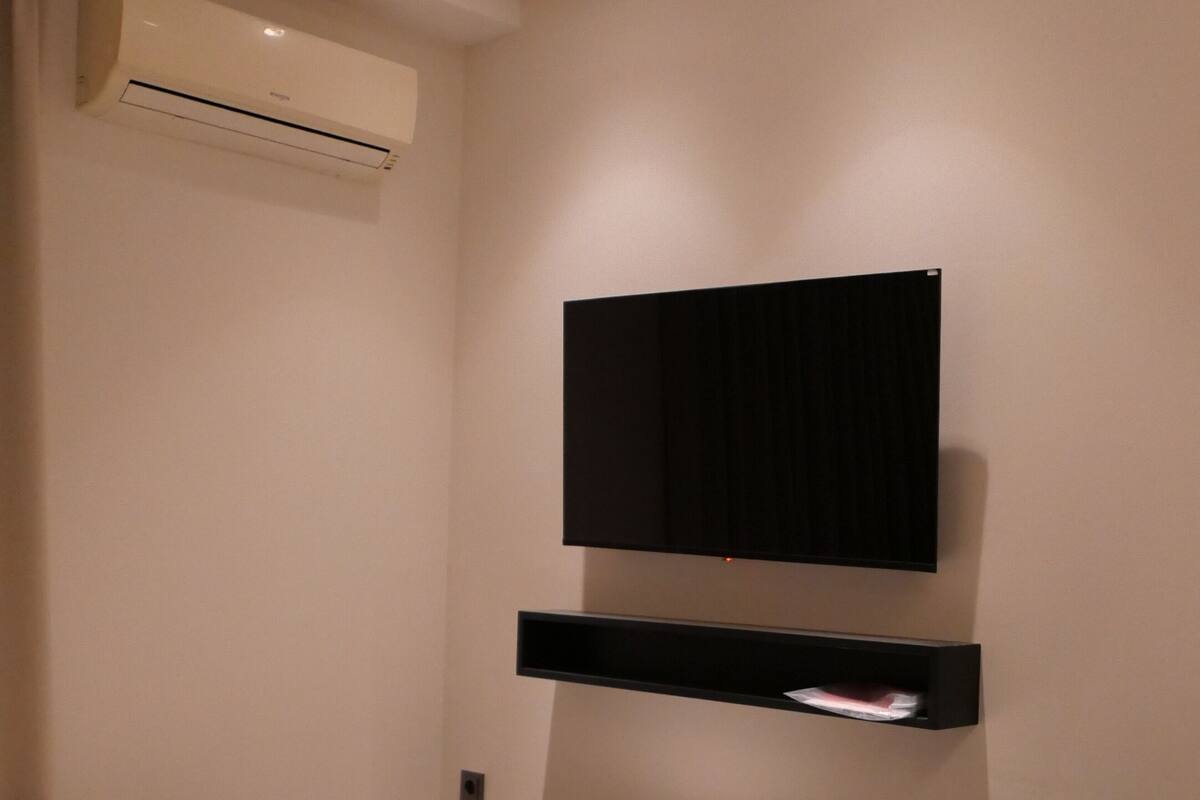 Quarto de hotel moderno com cama, uma mesinha redonda, televisão fixada na parede e ar condicionado integrado.