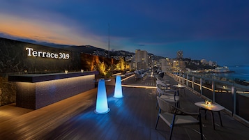 Terraço com placa indicando 'Terrace 30.9', piso de madeira, espreguiçadeiras e mesas com iluminação suave.