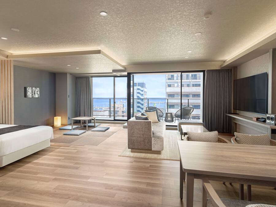 Quarto de hotel com cama, sofá, mesa de jantar, televisão e vista da cidade.